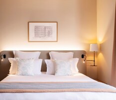Cama bem arrumada com lençóis brancos e almofadas estampadas com flores, um desenho arquitetônico emoldurado na parede e um abajur na mesa de cabeceira.
