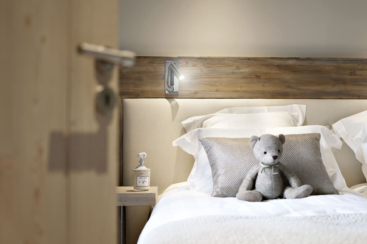 Cama bem arrumada com um urso de pelúcia, mesa de cabeceira com abajur e cabeceira de madeira.