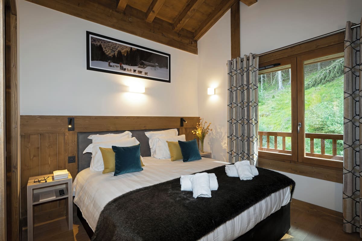 Quarto com cabeceira de madeira, uma cama grande com roupa de cama branca e várias almofadas, mesa de cabeceira com abajur e janela com vista para vegetação.