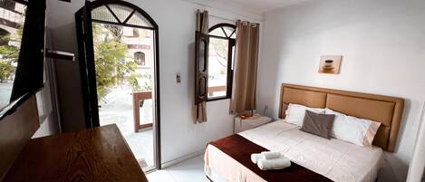 Quarto de hotel com cama, um banco de madeira, janela com cortinas e vista para o exterior.