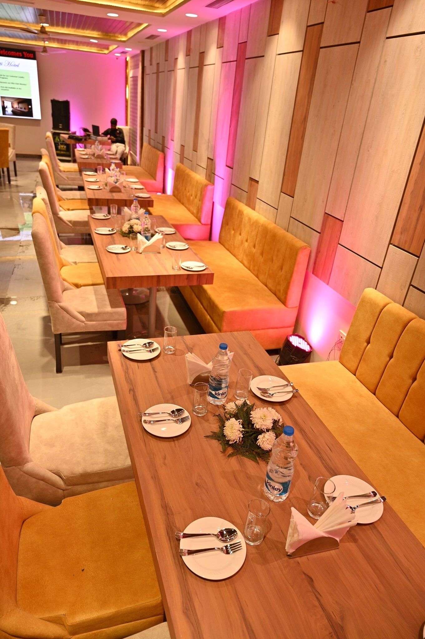 Uma sala de jantar com longas mesas de madeira dispostas para quatro pessoas, cada uma com um arranjo floral, garrafas de água e talheres.