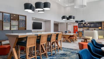 Um lobby de hotel moderno com área de bar, espaços de estar e uma cozinha visível ao fundo.