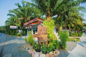Entrada do resort com placa de madeira, prédio com telhado vermelho e vegetação tropical.