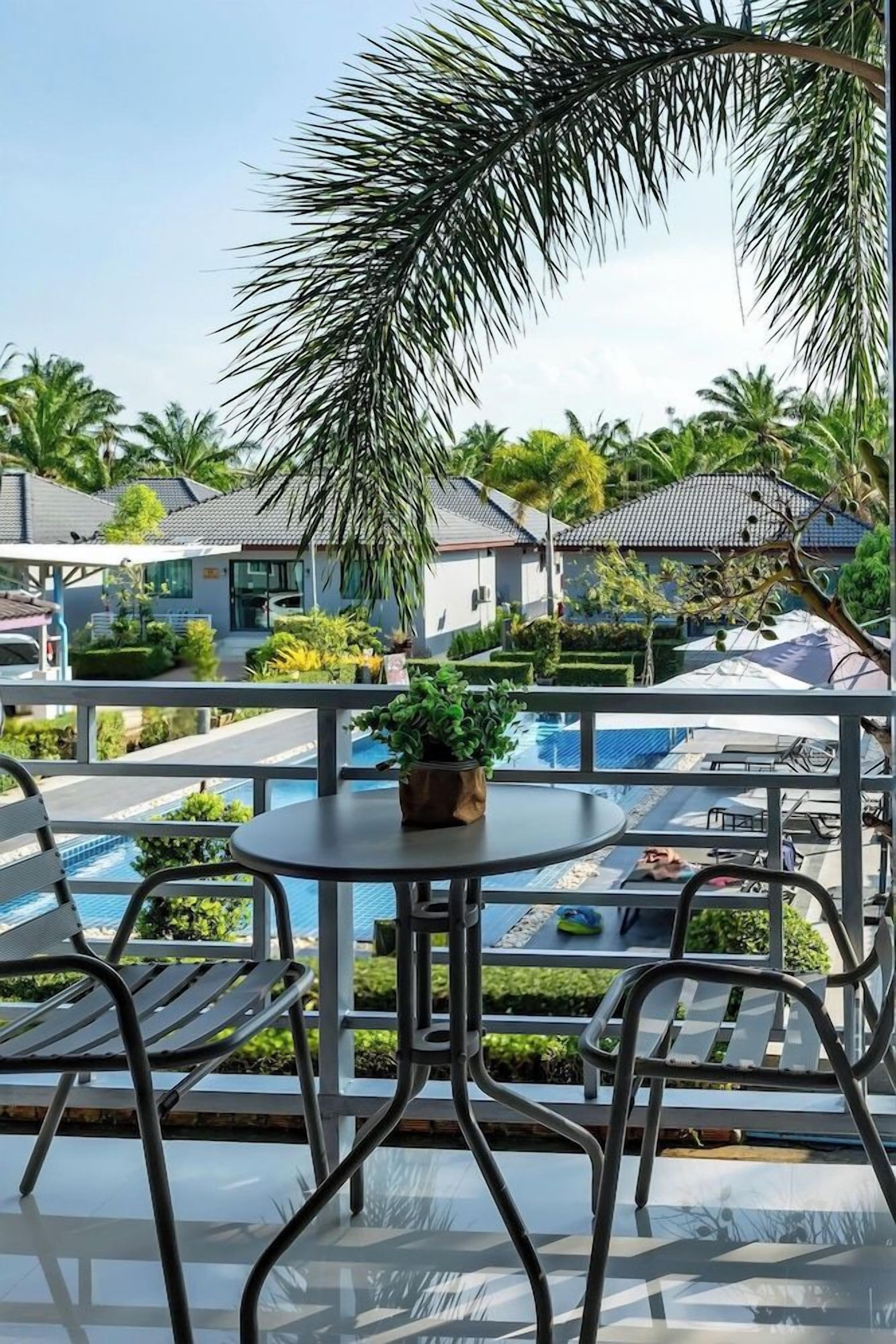 Varanda com mesa e cadeiras, com vista para uma piscina e área ajardinada com palmeiras e edifícios.