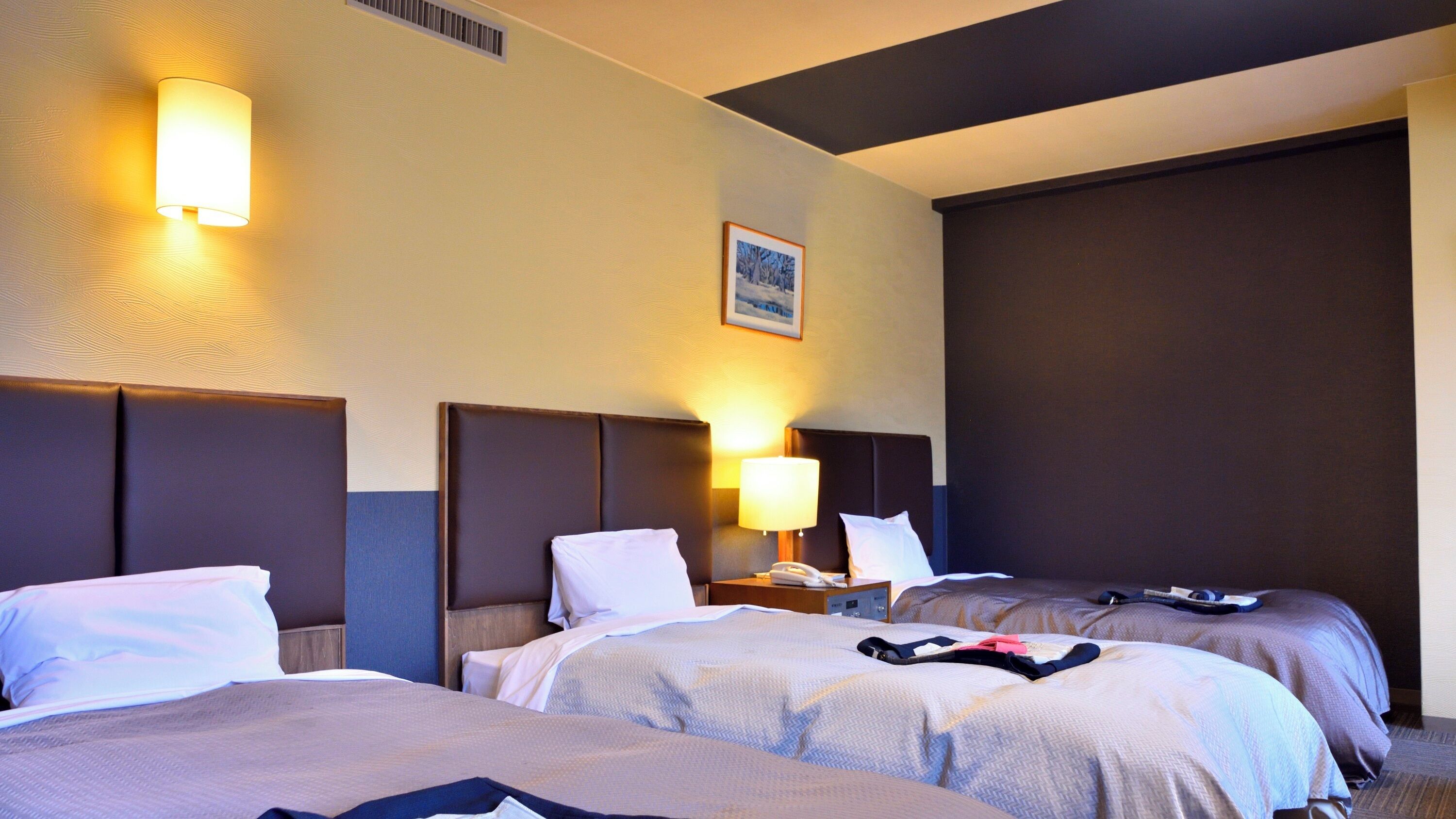 Quarto de hotel com duas camas, uma mesinha de cabeceira com abajur, um quadro pendurado na parede e uma janela grande.