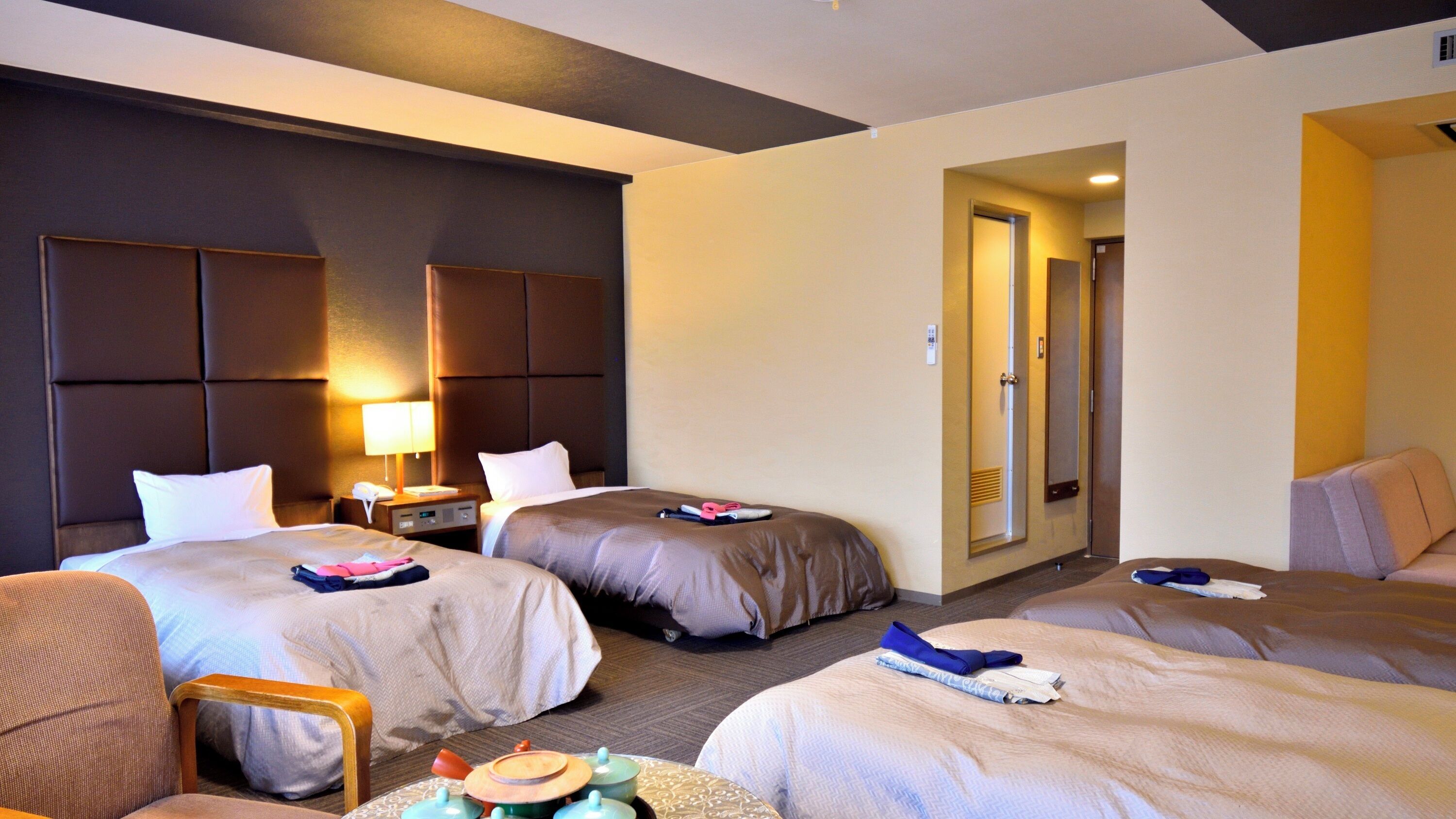 Quarto de hotel com três camas, uma cadeira, mesa de cabeceira com luminária e um espelho.