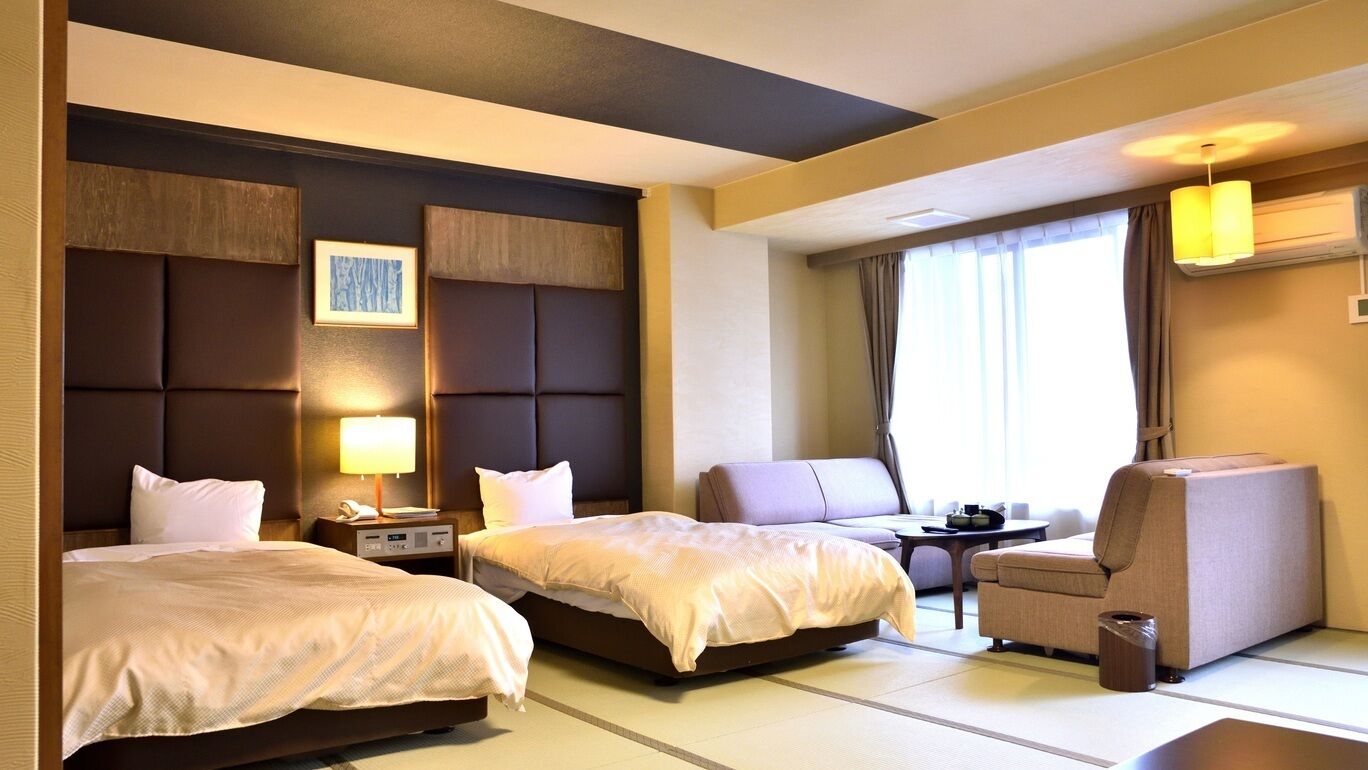 Quarto de hotel com duas camas, um sofá, uma poltrona, uma mesinha e um abajur.