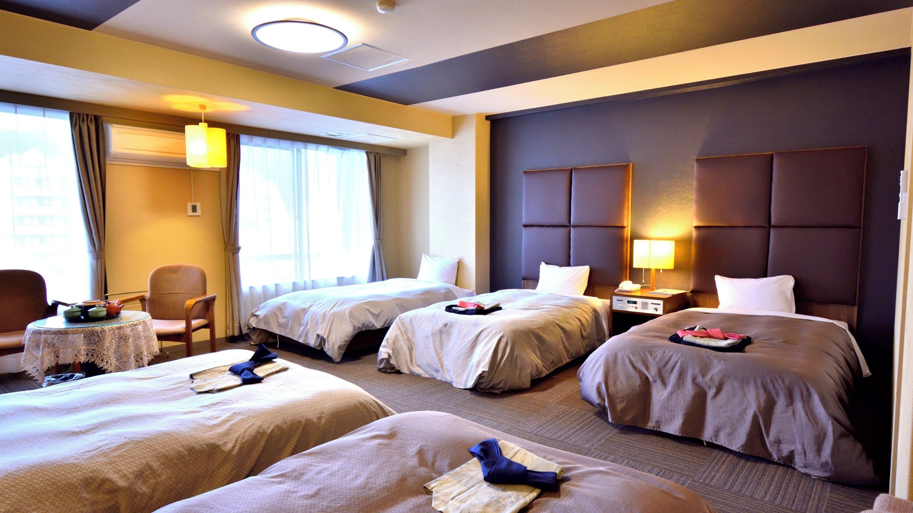 Quarto de hotel com duas camas, uma mesinha, uma cadeira e uma janela com cortinas.