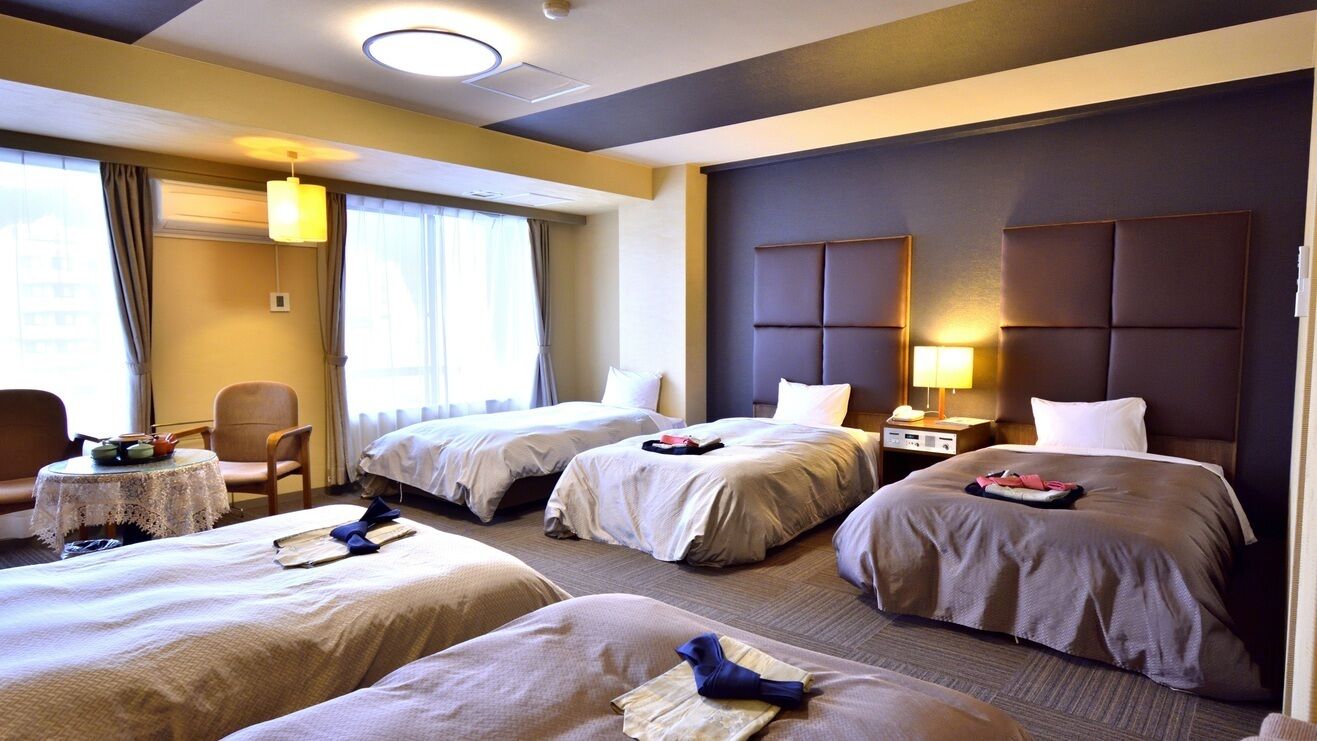 Quarto de hotel com três camas, uma mesinha com abajur e vista para a cidade.