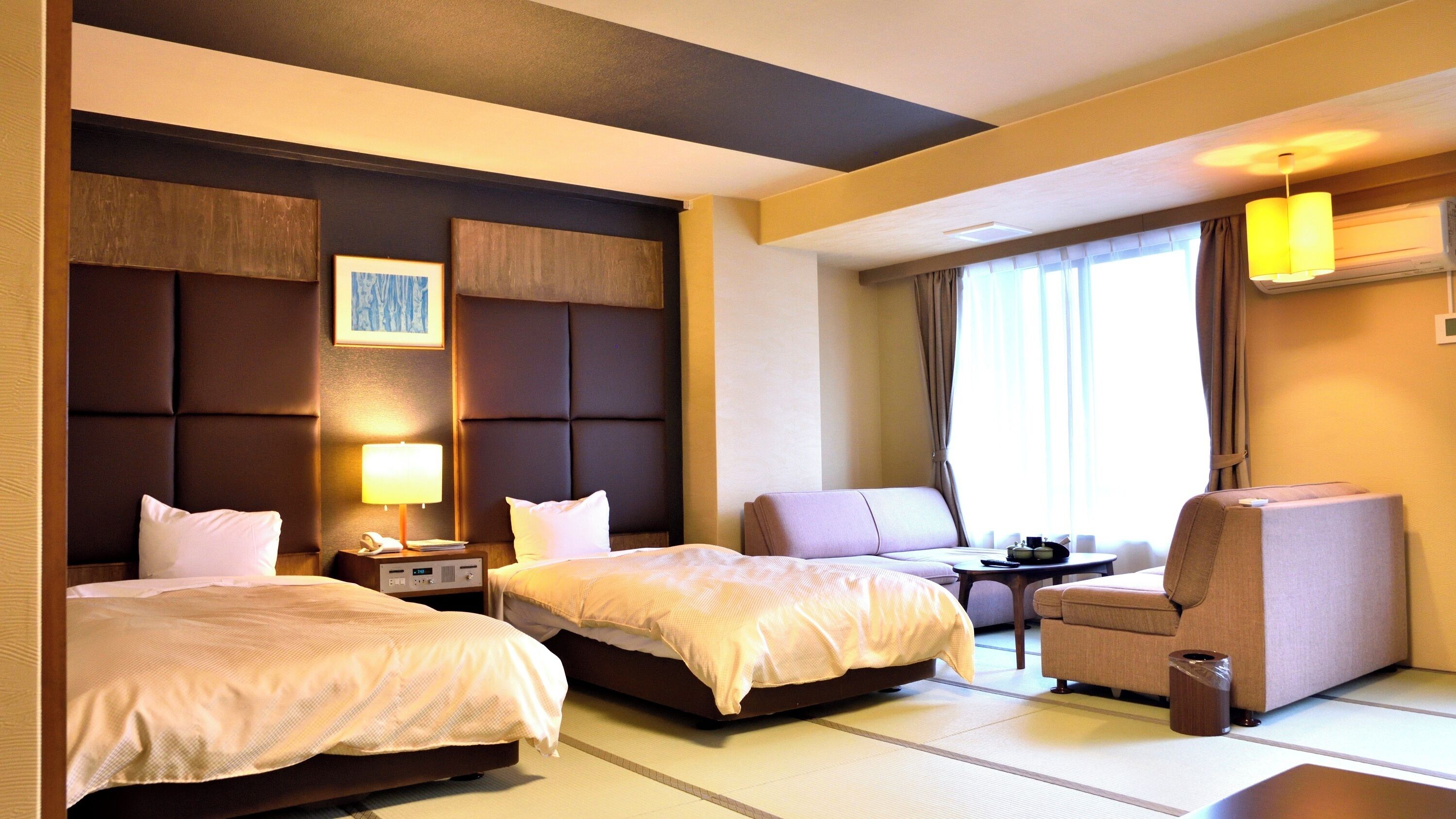 Quarto de hotel com duas camas, uma escrivaninha, uma cadeira, uma luminária e uma janela com cortinas.