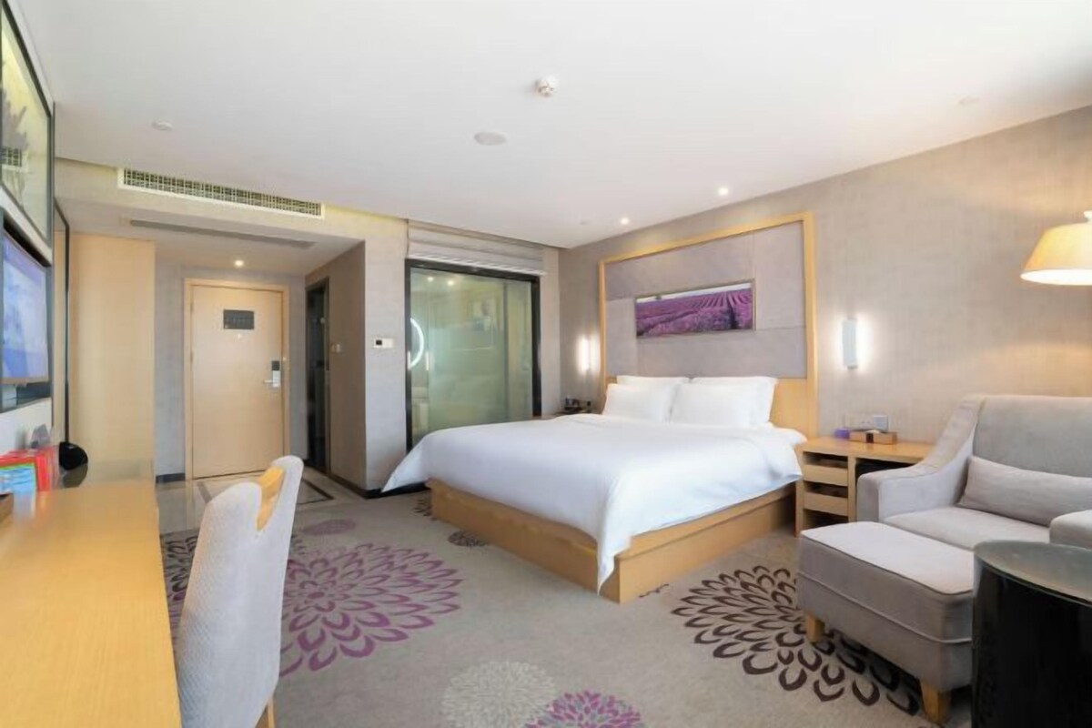 Quarto de hotel com cama, escrivaninha, poltrona, sofá e televisão presa na parede.