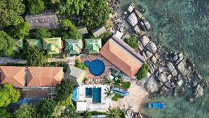 Vista aérea de um resort com vários edifícios, uma piscina central e uma área de praia rochosa.