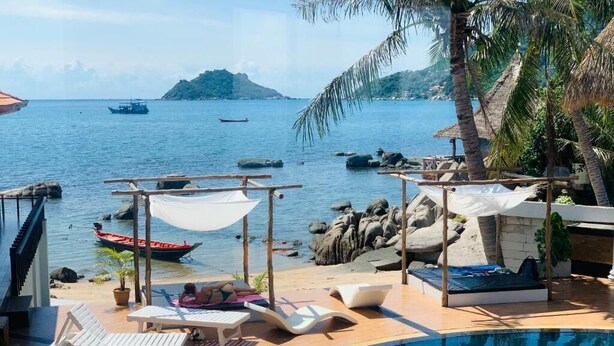 Uma área à beira da piscina com espreguiçadeiras, uma piscina pequena e vista para o mar com barcos.