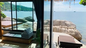 Quarto com uma grande janela com vista para a costa rochosa e o mar. Há uma cama com vista, uma mesa pequena e um banco.