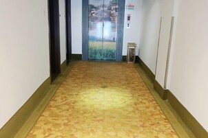 Corredor com carpete estampado, porta de elevador de vidro e uma pequena mesa com um aviso.