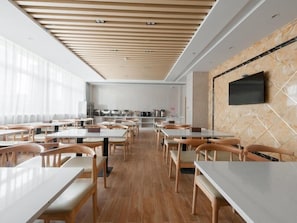 Uma sala de jantar moderna com mesas e cadeiras de madeira, cozinha ao fundo e uma TV de tela plana na parede.