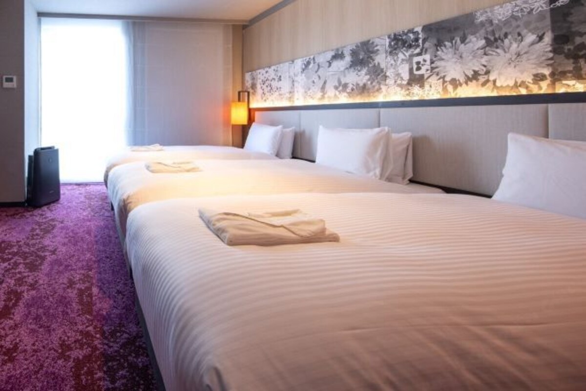 Quarto de hotel com duas camas, uma janela grande, um ornamento floral na parede e um abajur.