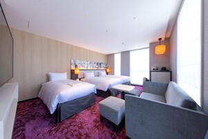 Quarto de hotel com duas camas, um sofá cinzento, uma mesinha e uma janela grande.