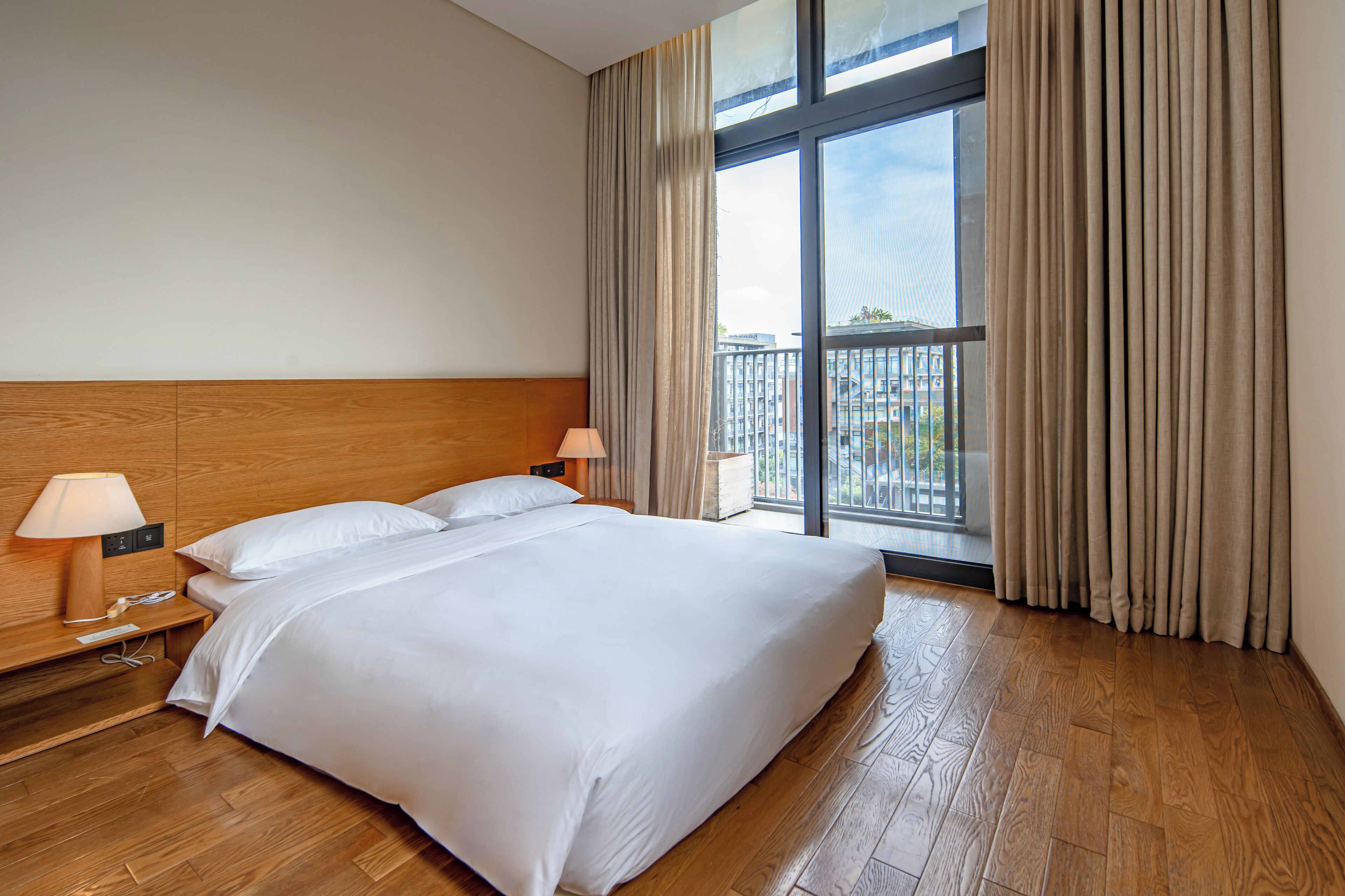Quarto de hotel com uma cama grande, cabeceira de madeira, duas luminárias de apoio e vista para uma varanda através de janelas do chão ao teto.