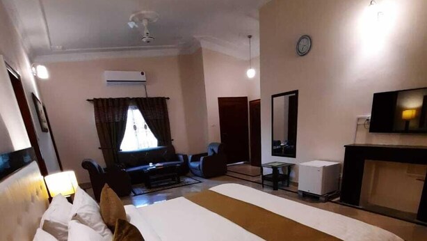 Quarto de hotel com uma cama grande, uma escrivaninha com abajur, uma televisão e um relógio na parede.
