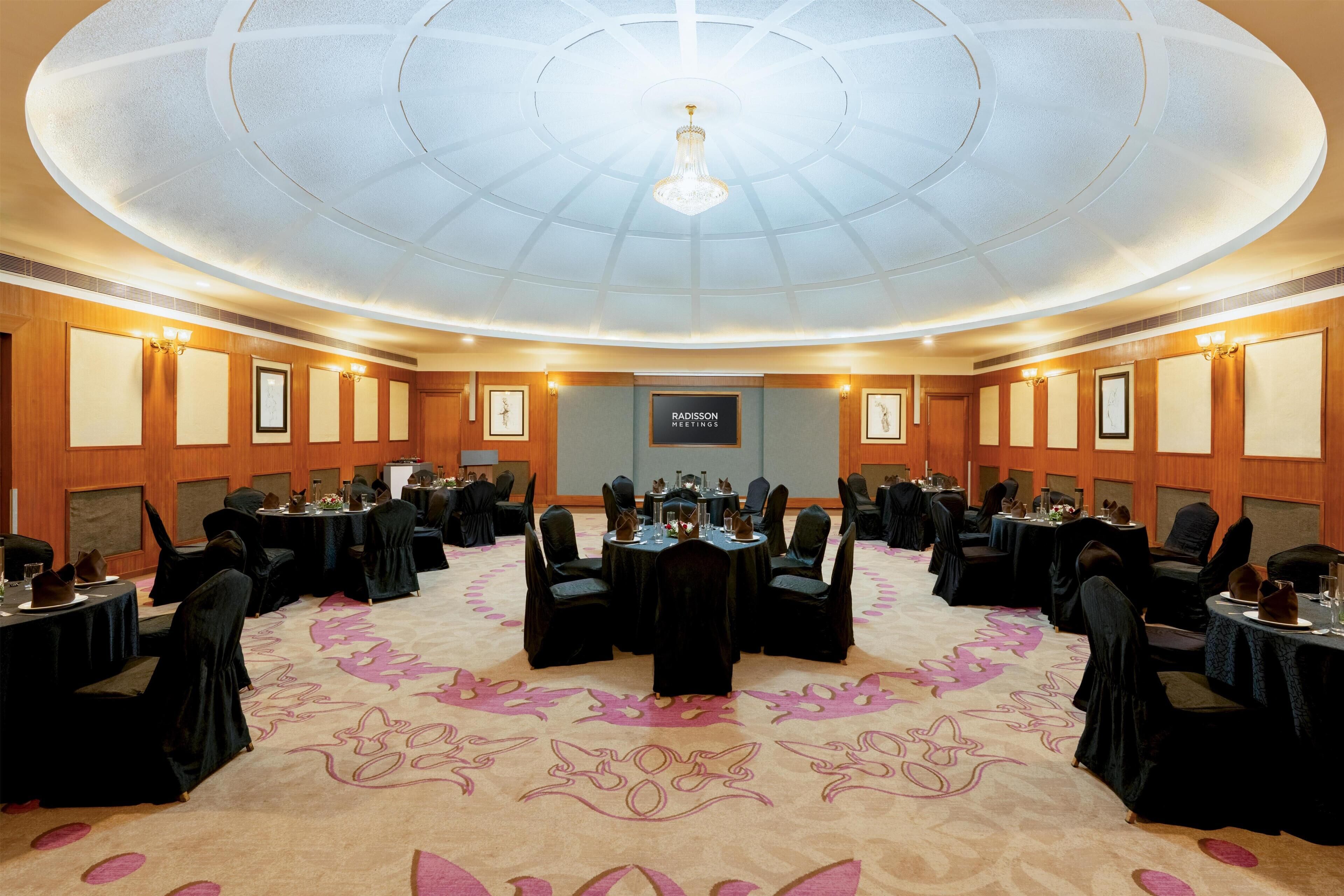 Uma sala de conferências com mesas redondas, cadeiras pretas e um teto com luminária central.
