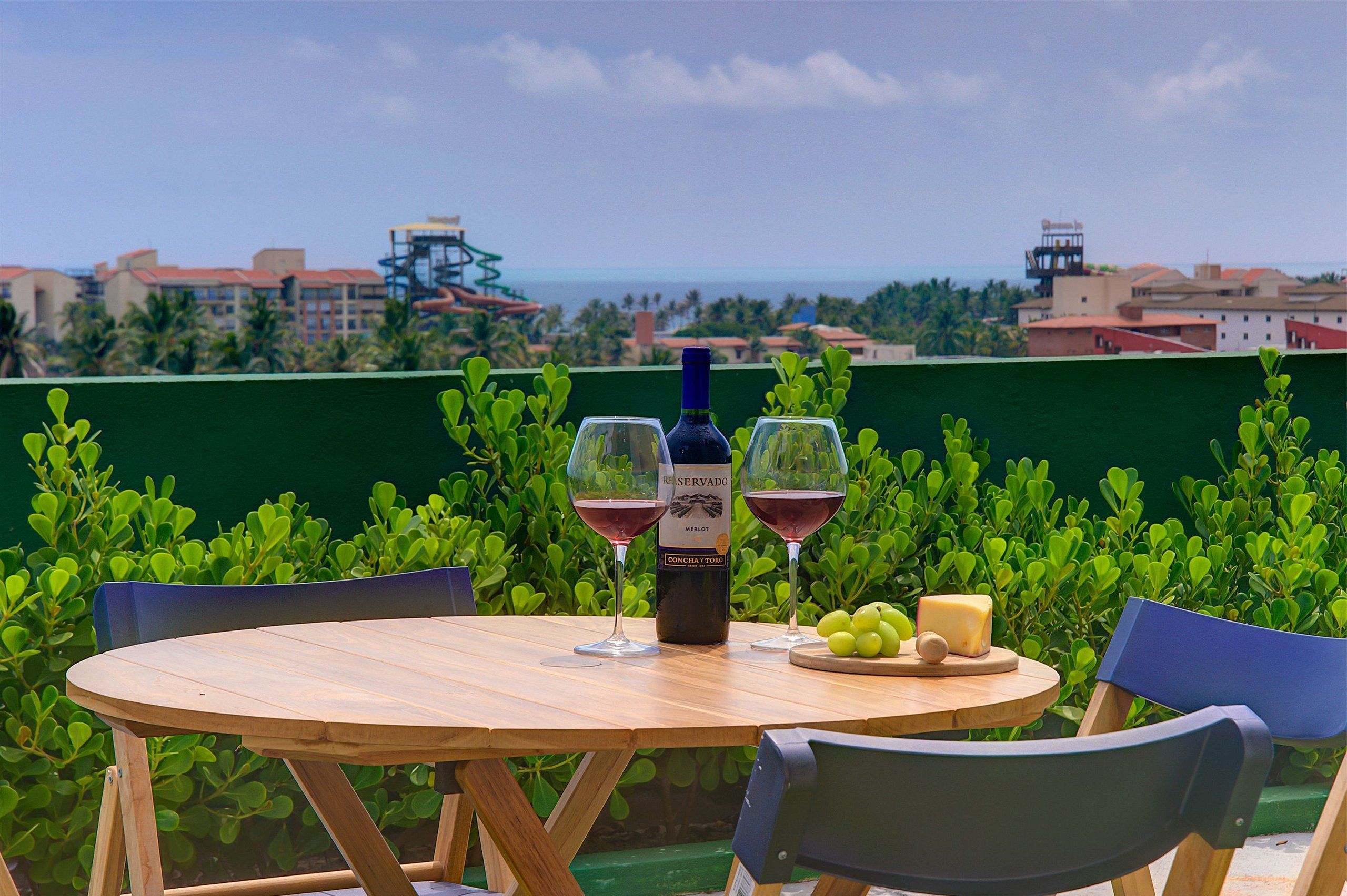 Mesa de madeira redonda com duas taças de vinho e uma garrafa, um prato com queijo e uvas, cadeiras, e um fundo com um hedge verde e edifícios com vista para o mar.