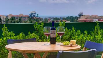 Mesa de madeira redonda com duas taças de vinho e uma garrafa, um prato com queijo e uvas, cadeiras, e um fundo com um hedge verde e edifícios com vista para o mar.