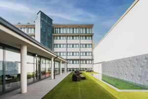 Um edifício moderno com área externa coberta, grama sintética e mesas para o ar livre.