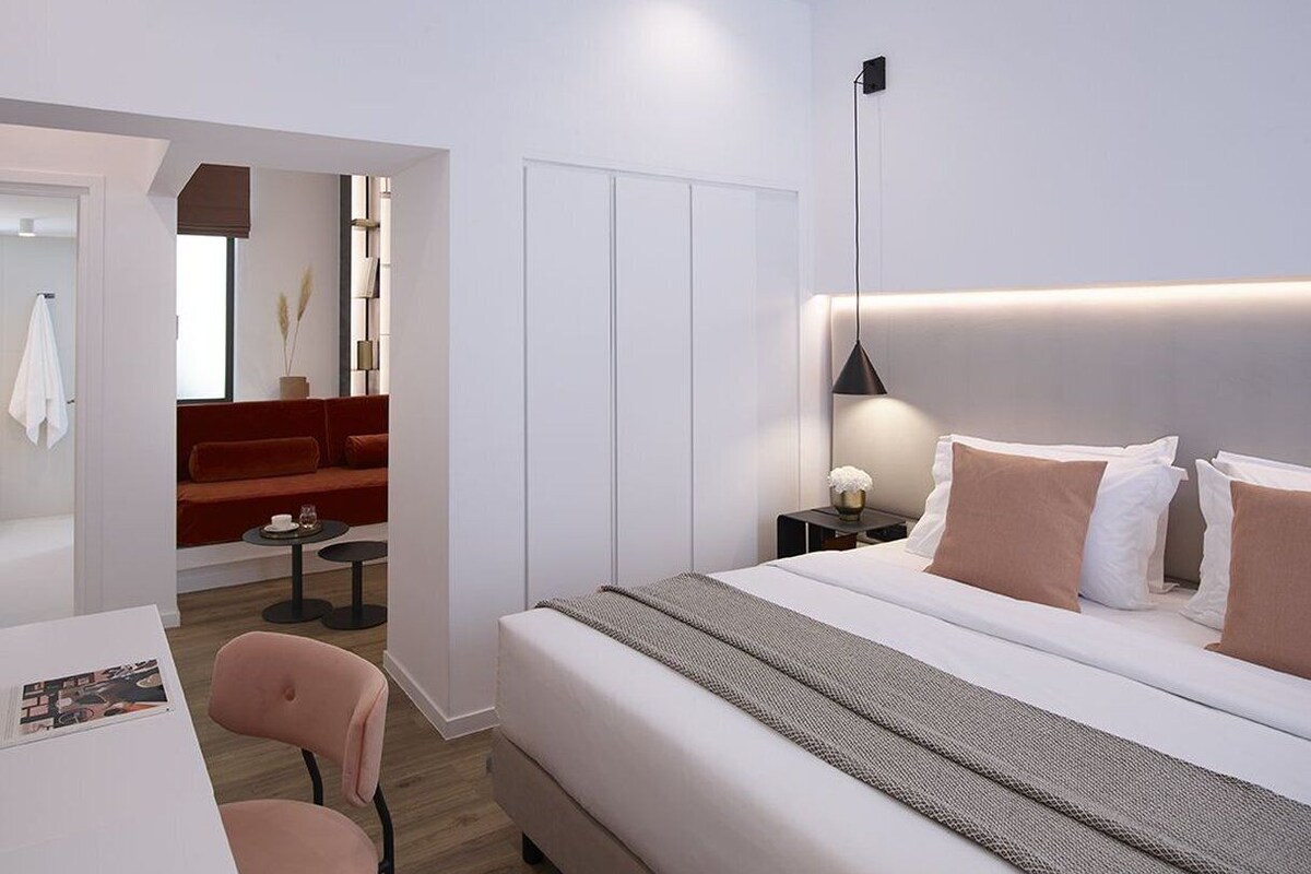 Quarto de hotel moderno com uma cama grande, mesas de cabeceira, uma escrivaninha e um sofá vermelho ao fundo.
