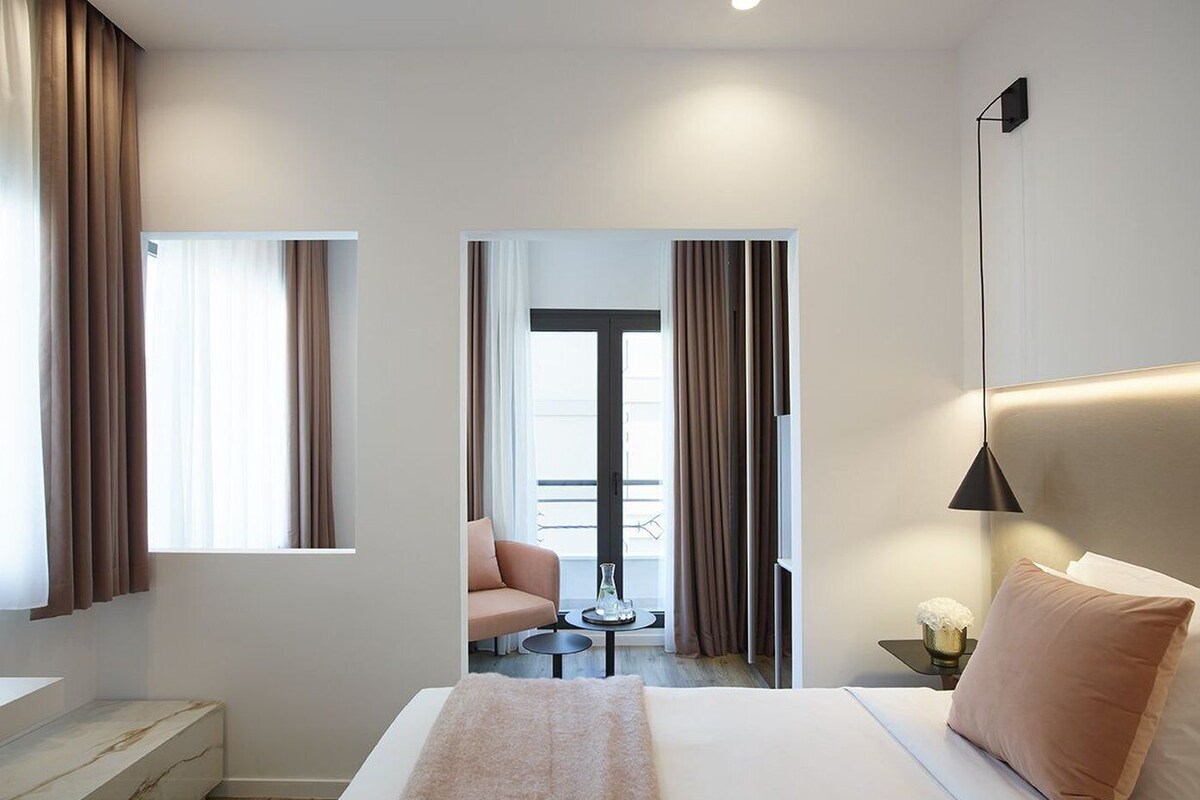 Quarto de hotel com cama, mesa de cabeceira, luminária, janela com cortinas e vista exterior.