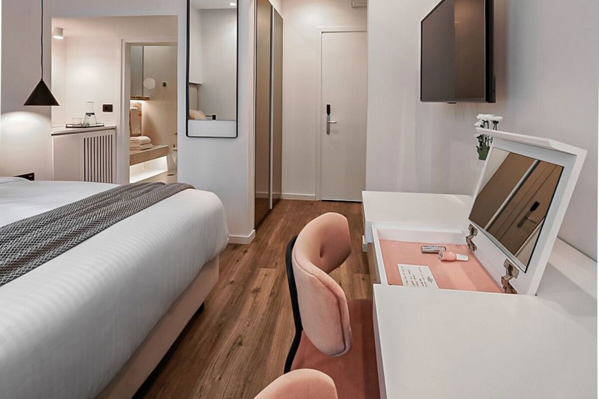 Quarto de hotel moderno com cama, escrivaninha, cadeira, televisão e espelho.