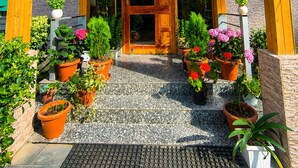 Entrada de um prédio de madeira com varanda coberta, plantas em vasos e muro de pedra.