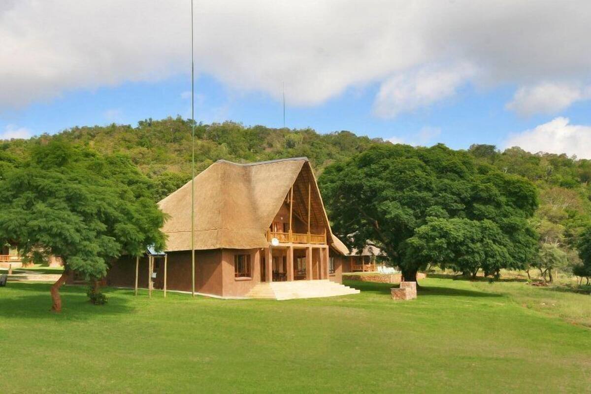 Um edifício com telhado de palha e um deque de madeira, rodeado por vegetação exuberante e colinas.