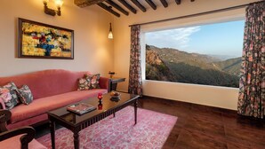 Quarto com sofá, mesa de centro, um quadro e vista para uma paisagem montanhosa através de uma grande janela.