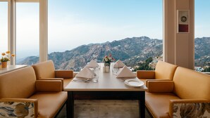 Uma área de refeições com uma mesa posta para dois, grandes janelas com vista para as montanhas, e teto com vigas e luminárias aparentes.