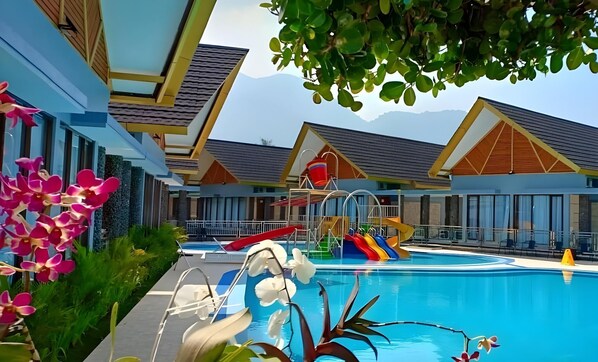 Piscina com escorregador e área de recreação, rodeada por edifícios com grandes janelas e céu claro.