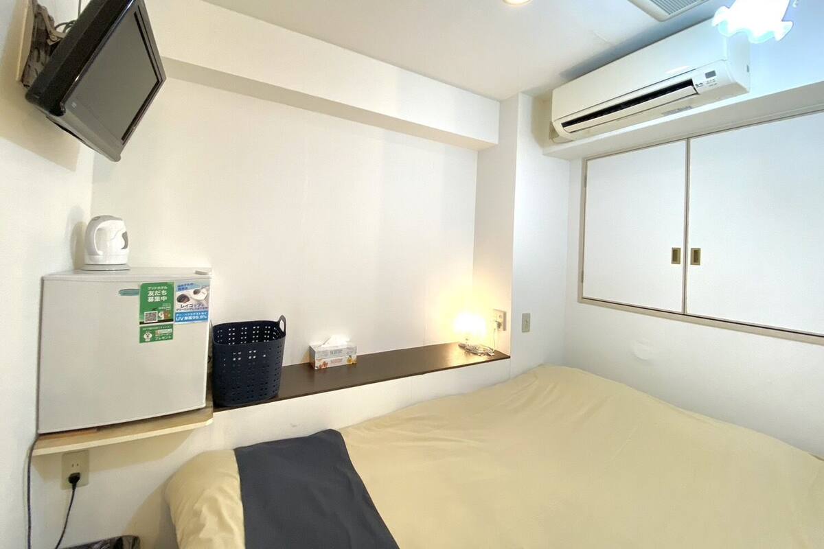 Quarto de hotel com cama, uma pequena escrivaninha com luminária, televisão, frigorífico e ar condicionado.
