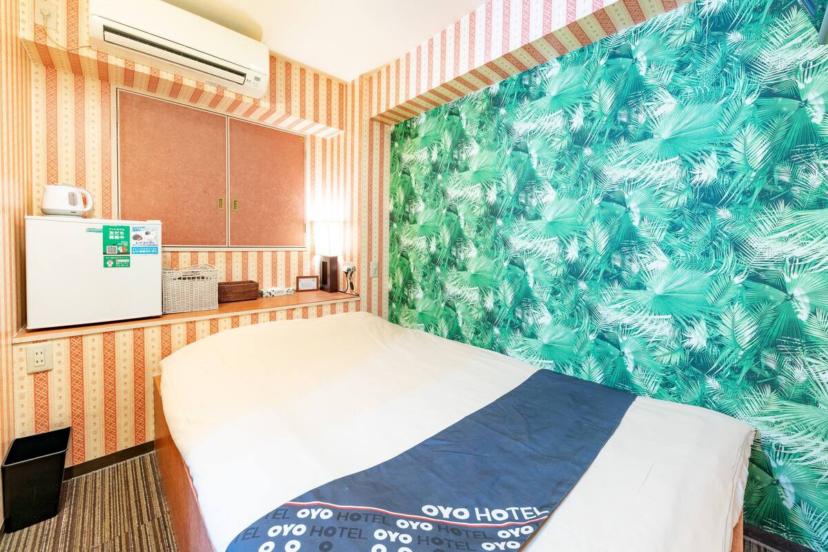 Quarto de hotel com cama, pequeno espaço de cozinha, espelho e parede com padrão de folhagem tropical.