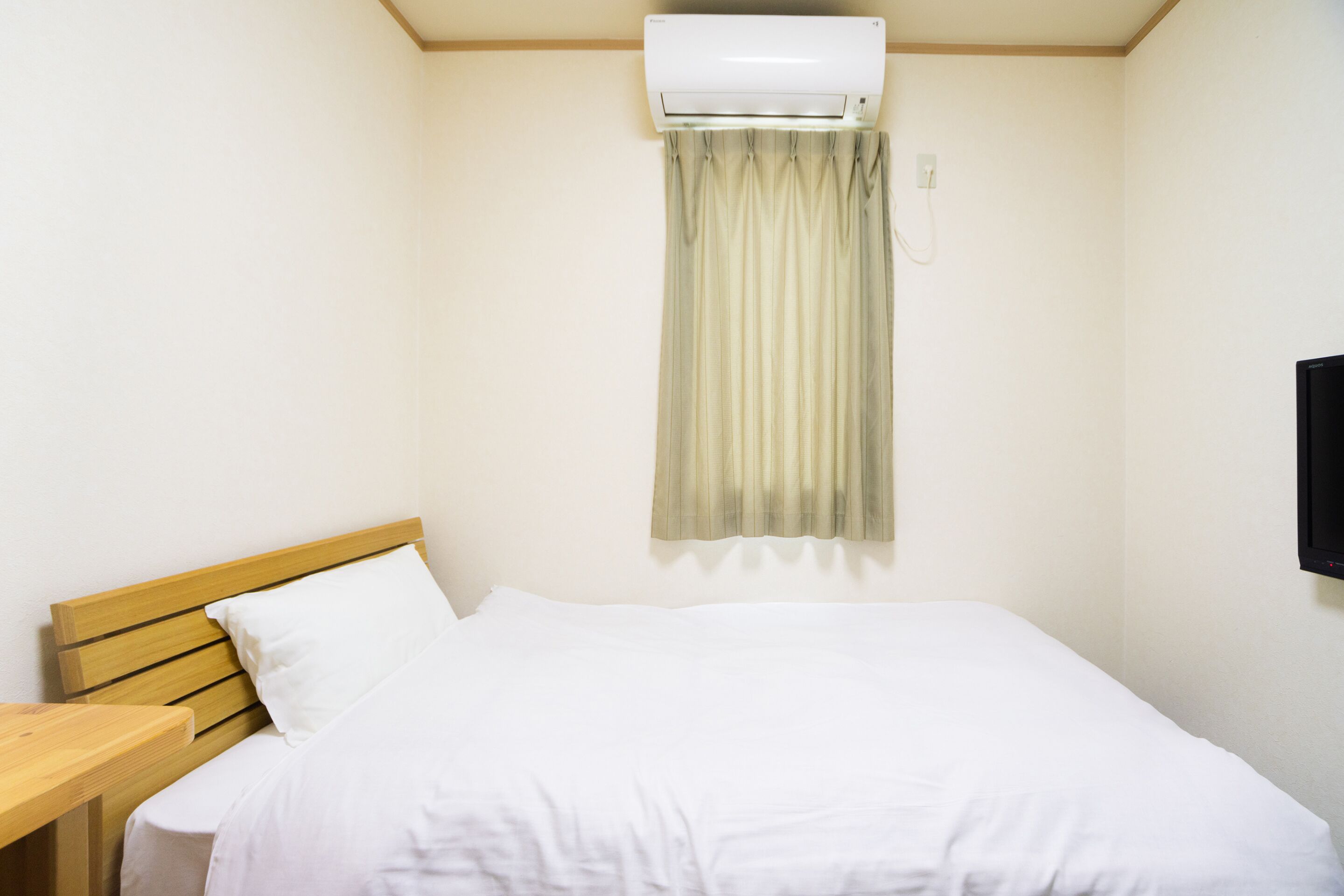 Quarto de hotel com uma cama de solteiro, cabeceira de madeira, colchão branco, mesa de cabeceira de madeira, ar condicionado fixado na parede e televisão na parede.