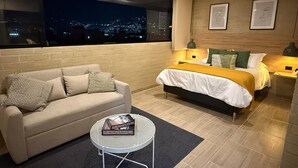 Quarto moderno com vista para a cidade, um sofá, uma cama com manta amarela e duas luminárias de cabeceira.
