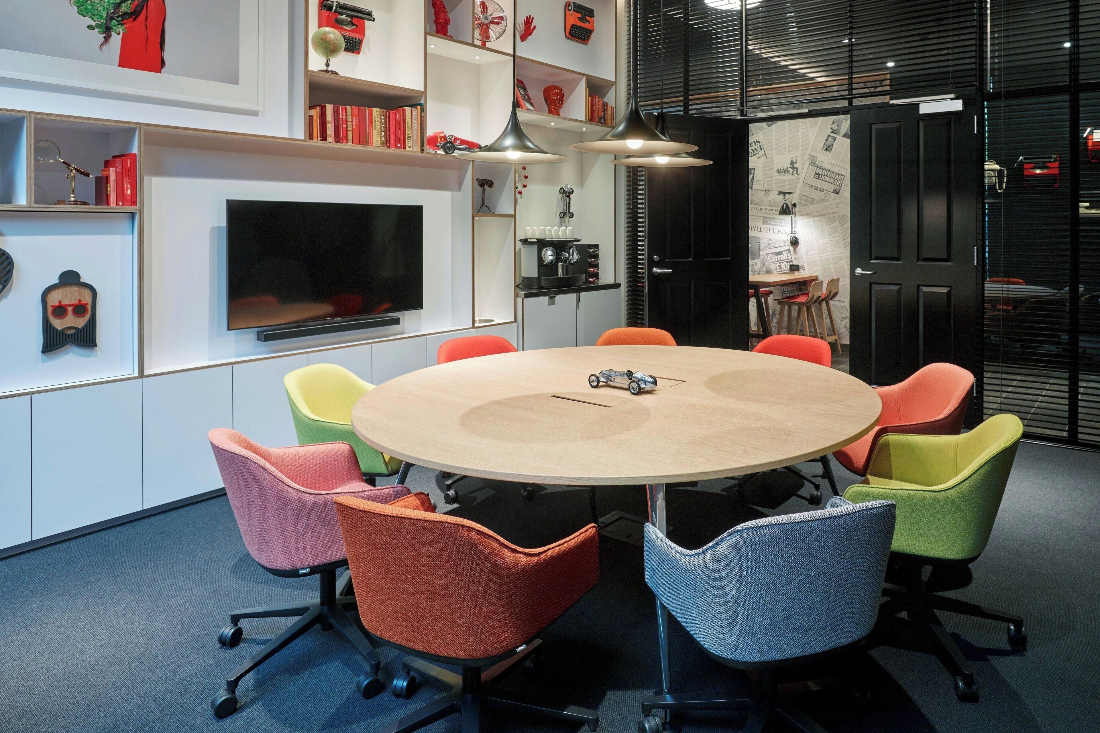 Uma sala de conferências com uma mesa redonda, cadeiras coloridas, uma televisão, estantes com livros e objetos decorativos, e uma porta com parede coberta de jornal.