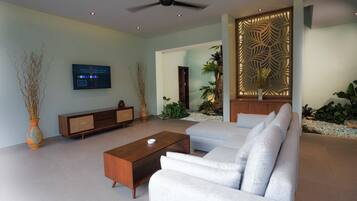 Uma sala de estar moderna com um sofá branco grande, uma mesa de centro de madeira, uma TV fixada na parede e uma parede decorativa com plantas.
