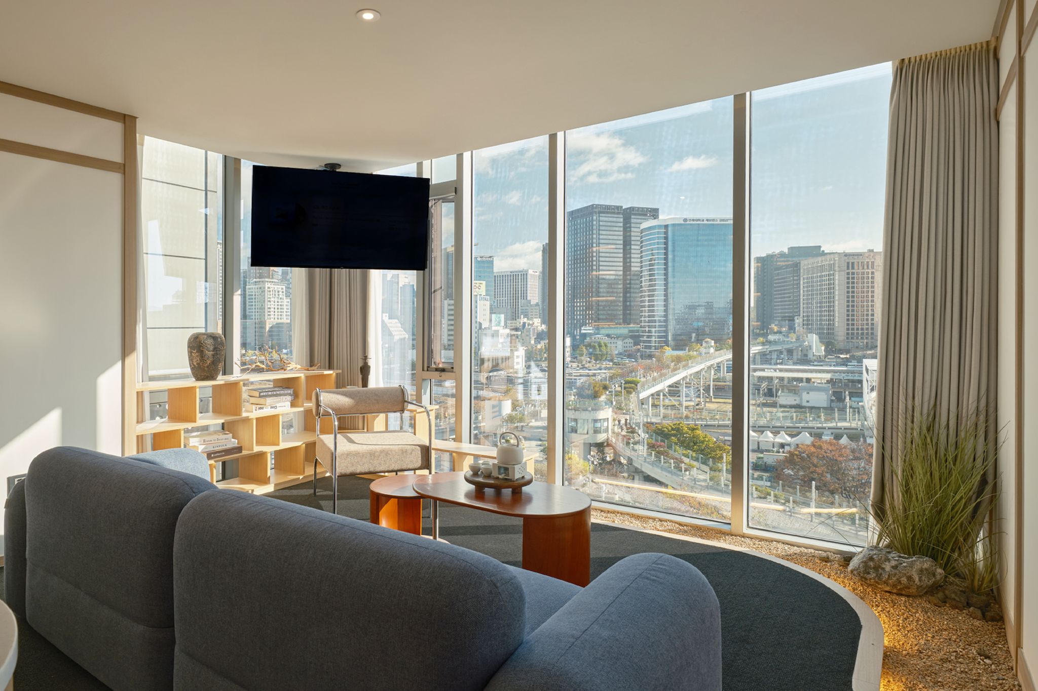 Sala de estar moderna com sofá, mesa de centro e televisão. Vista da cidade através de amplas janelas.