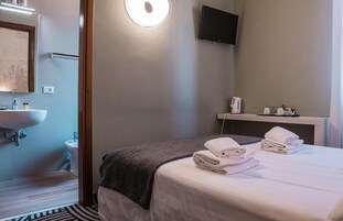 Quarto de hotel com cama, mesa de cabeceira com produtos de higiene pessoal, televisão na parede e banheiro com pia e vaso sanitário.