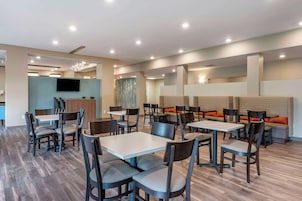 Uma sala de jantar moderna com várias mesas e cadeiras, uma TV de tela plana na parede e um painel de madeira com almofadas laranja.