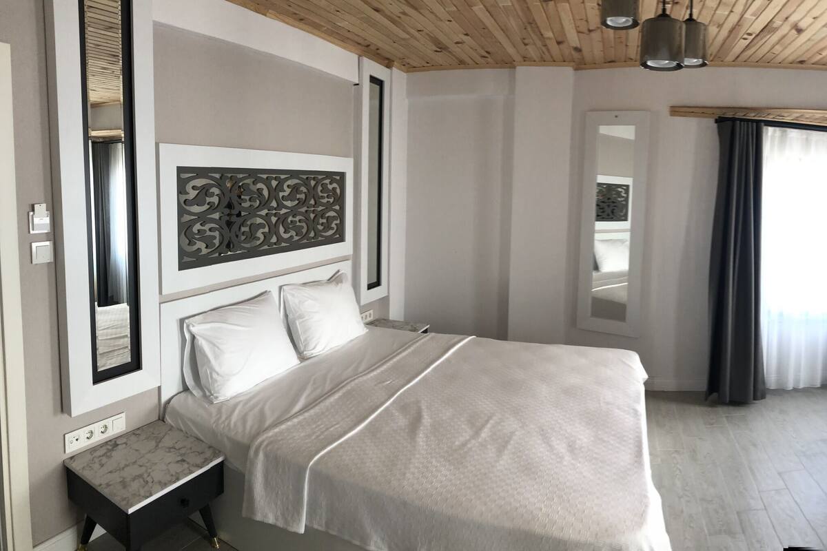 Quarto com cama, mesa de cabeceira, espelho, televisão e janela com cortinas.