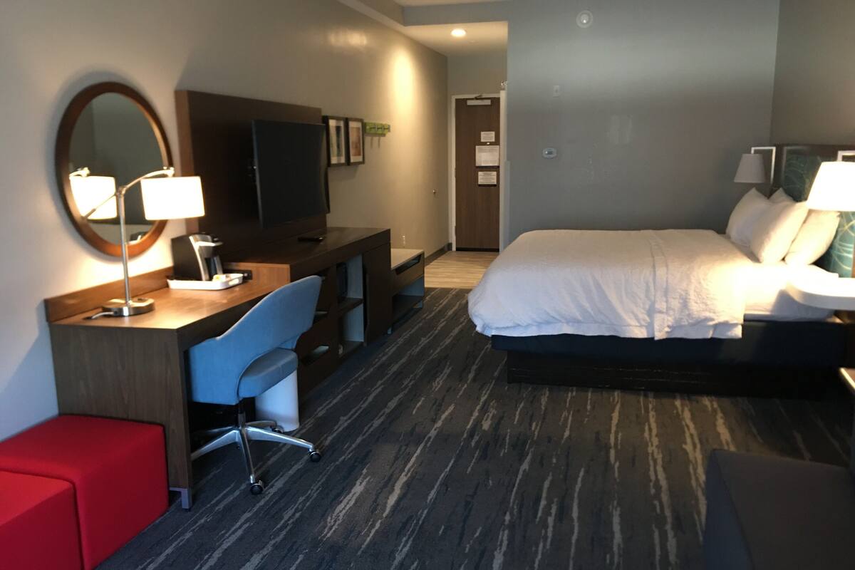 Quarto de hotel com cama, escrivaninha, cadeira e televisão.