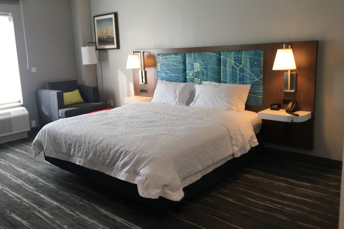 Quarto de hotel com uma cama grande, duas mesas de cabeceira com abajures, uma cadeira, um quadro na parede e uma janela.