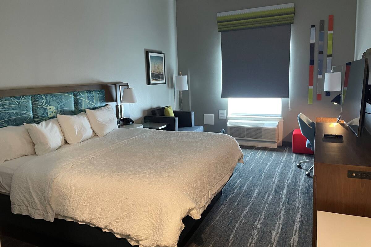 Quarto de hotel com uma cama grande, uma escrivaninha com computador, uma cadeira, uma TV e uma janela com persianas.