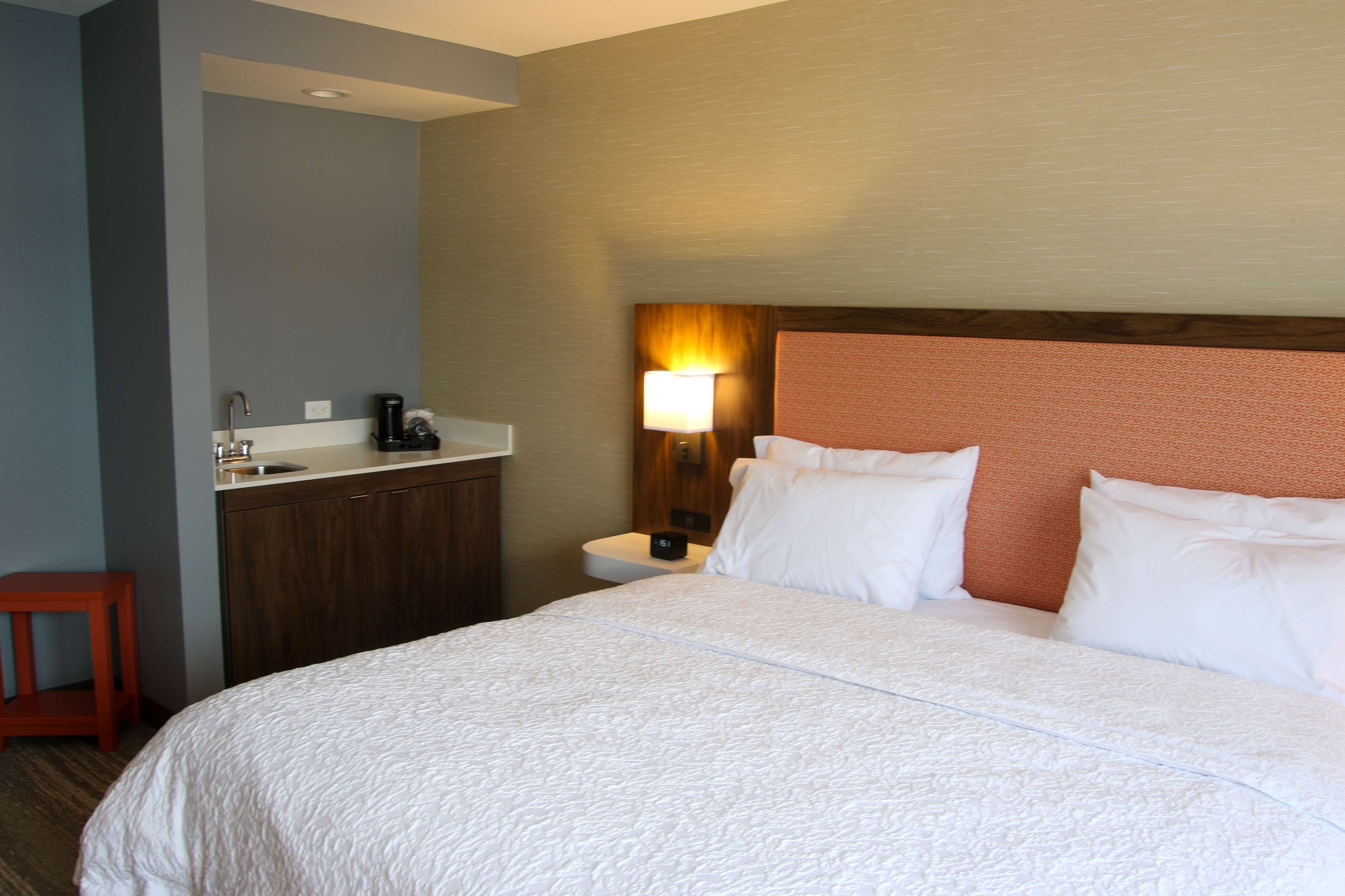 Quarto de hotel com uma cama grande, uma mesa de cabeceira com abajur, uma pequena escrivaninha com cafeteira e um armário de madeira.